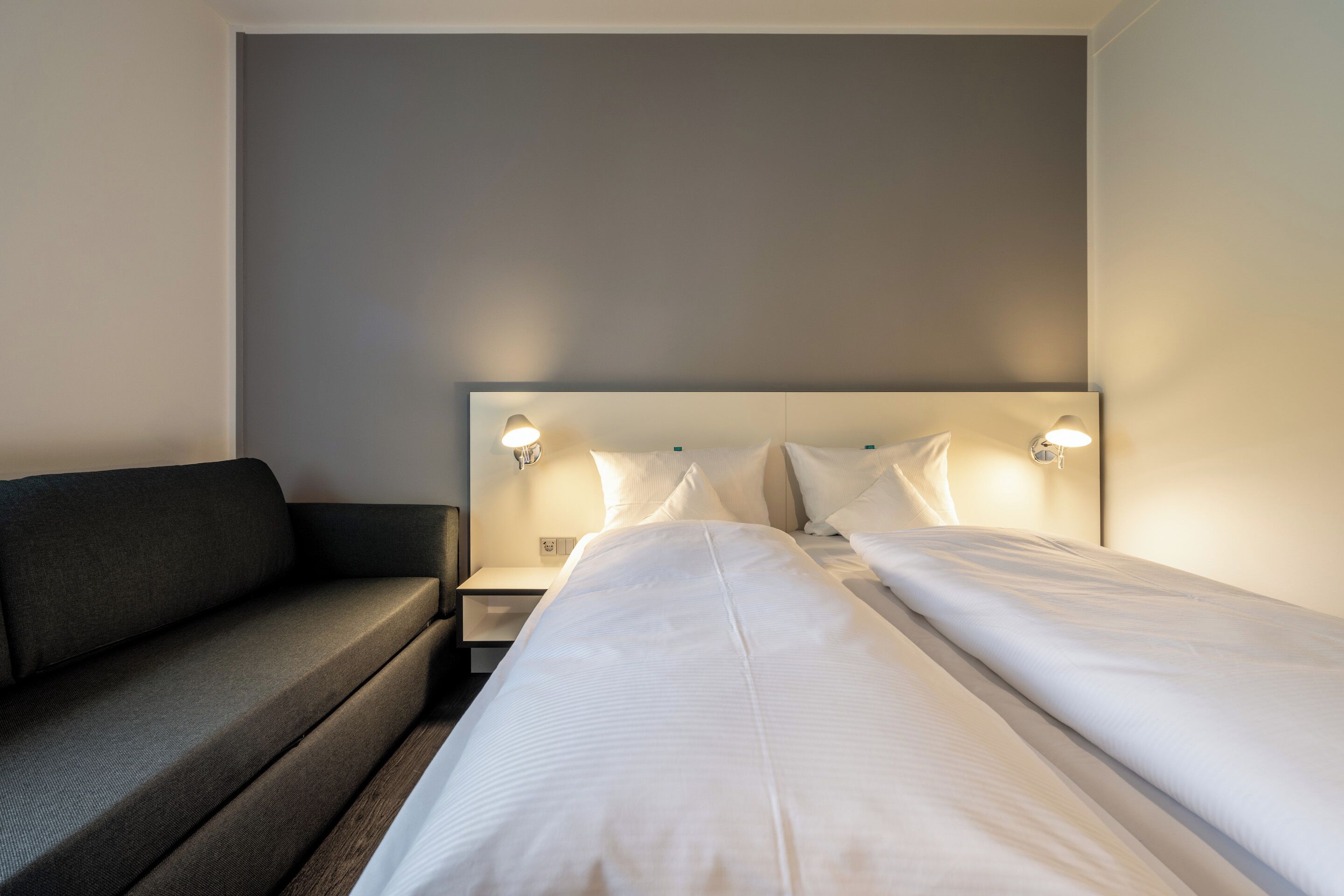 Quarto de hotel com uma cama, um sofá e duas luminárias de cabeceira.