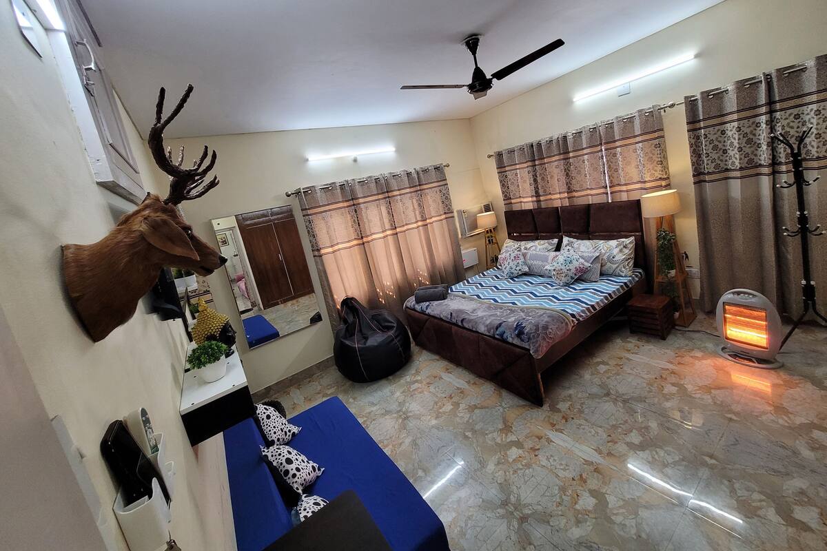 Quarto com cama, ventoinha no teto, aquecedor, espelho, cortinas, cabeça de veado montada na parede e um sofá azul.