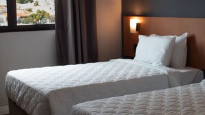 Quarto de hotel com duas camas, uma janela grande com cortinas, uma luminária na parede e vista para a paisagem urbana.