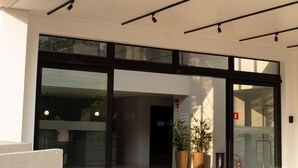 Entrada de um edifício moderno com grandes portas de vidro, plantas em vasos e um extintor próximo à entrada.