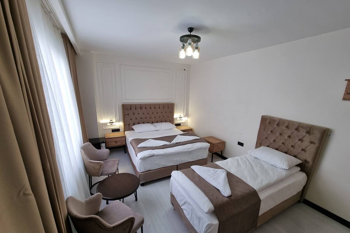 Quarto de hotel com duas camas, uma cadeira, uma mesinha e um candeeiro no teto.