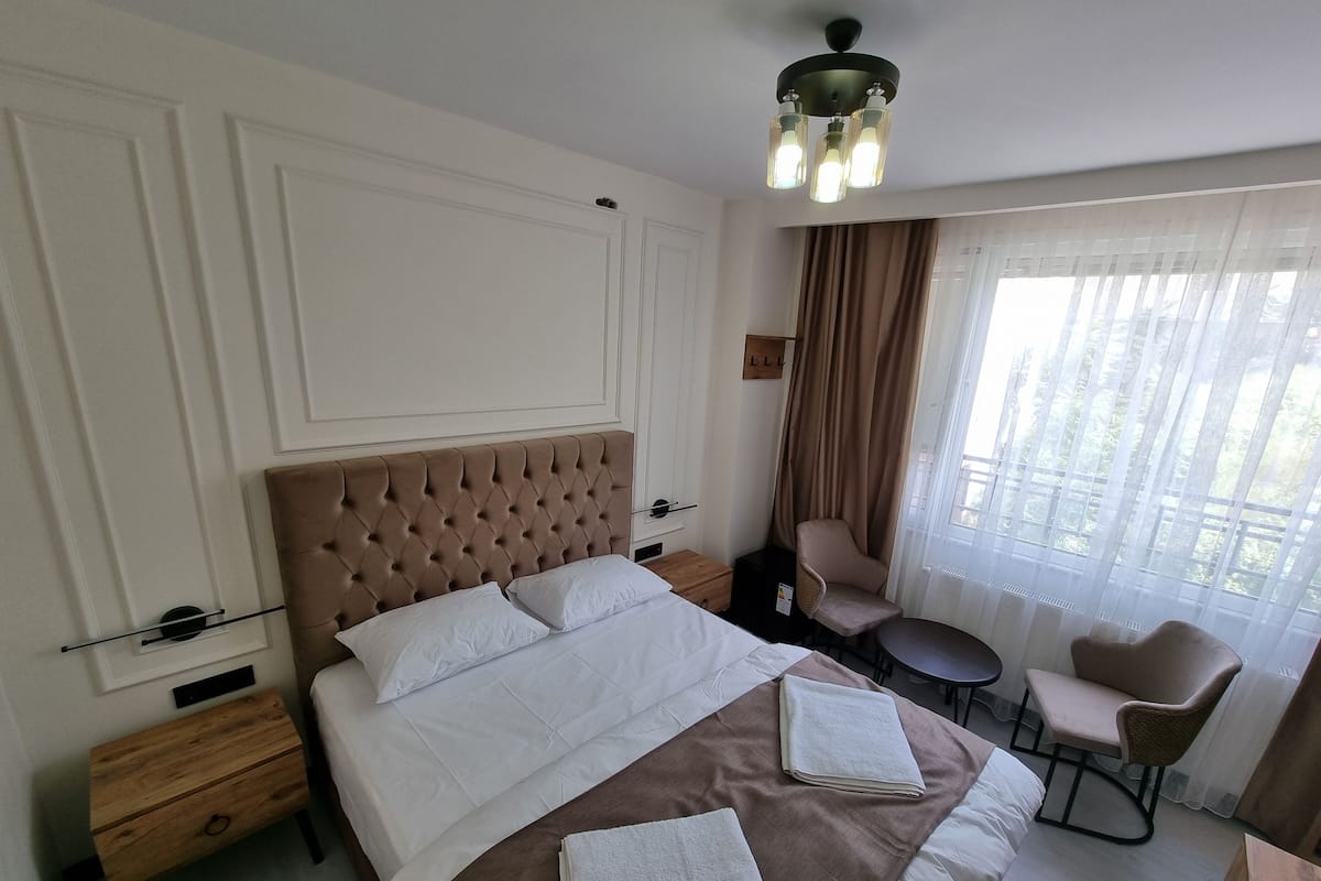Quarto de hotel com uma cama grande, uma mesinha de cabeceira de madeira, uma cadeira, uma pequena mesa e uma janela com cortinas.