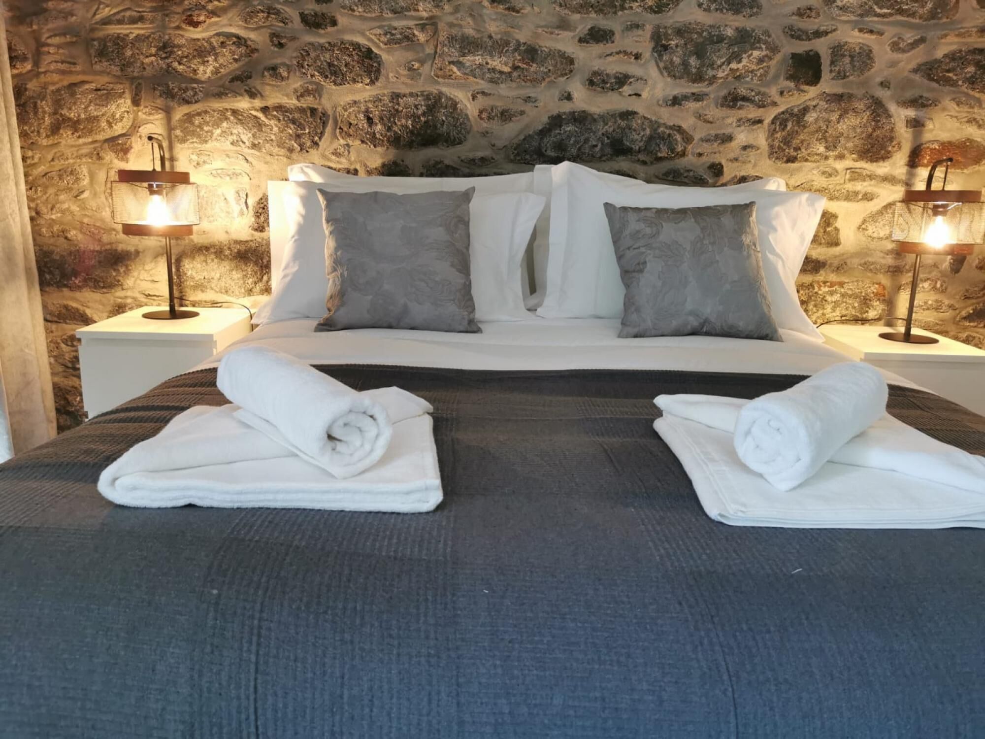 Uma cama com parede de pedra ao fundo, duas mesas de cabeceira e duas luminárias.