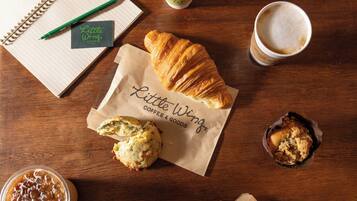 Uma mesa de madeira com um caderno, uma caneta, um croissant, um muffin e uma xícara de café.