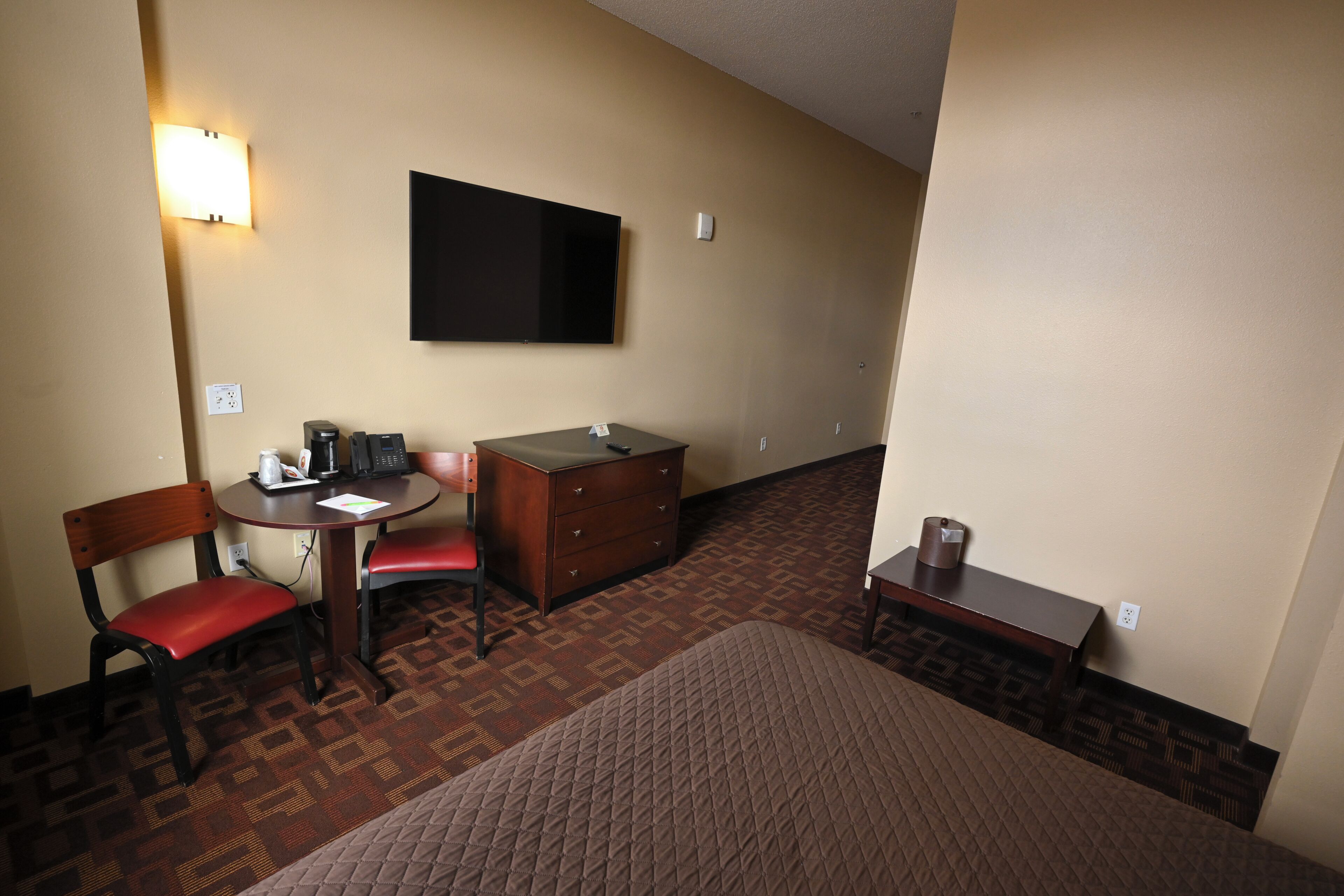 Quarto de hotel com uma cama, uma escrivaninha com cadeira, uma televisão na parede e um tapete estampado.