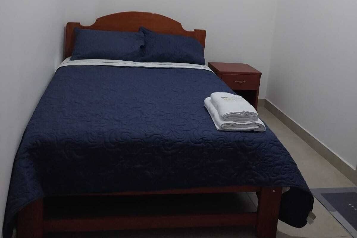 Quarto individual com uma cama de solteiro, cabeceira de madeira, colcha azul, travesseiro branco, uma pequena mesa de cabeceira e uma toalha dobrada no chão.