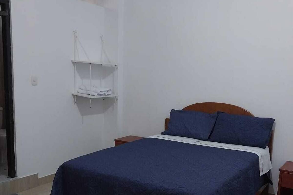 Quarto com cama, cabeceira de madeira, paredes brancas e uma pequena mesa de cabeceira.