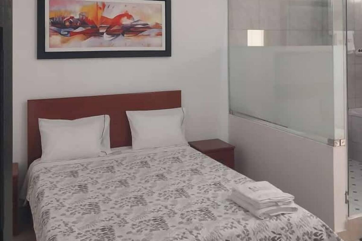 Quarto de hotel com cama, mesa de cabeceira, box de chuveiro de vidro e um quadro na parede.