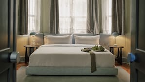 Uma cama bem arrumada com manta e uma planta, duas mesas de cabeceira com abajures, e uma janela com cortinas.