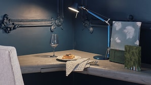 Um quarto escuro com uma escrivaninha, uma cadeira, um abajur, um copo de vinho e um prato de comida.