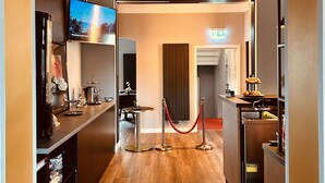 Um hall de hotel moderno com piso de madeira, uma televisão fixada na parede e uma cozinha visível através de uma porta aberta.