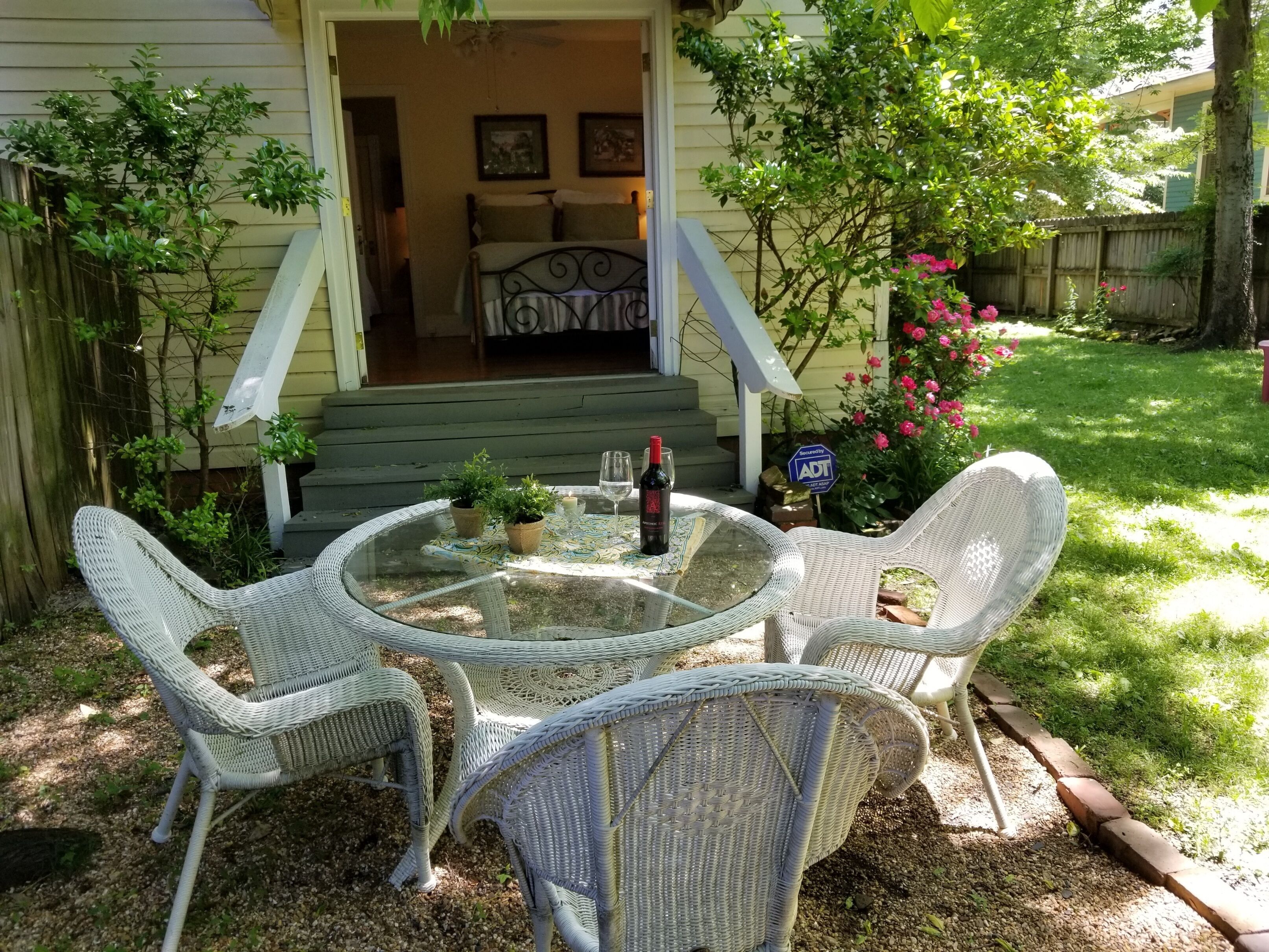 Mesa com tampo de vidro e cadeiras de vime num quintal com uma casa ao fundo.