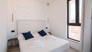 Cama bem arrumada com almofadas e toalhas, uma mesa de cabeceira e uma janela com vista para edifícios.