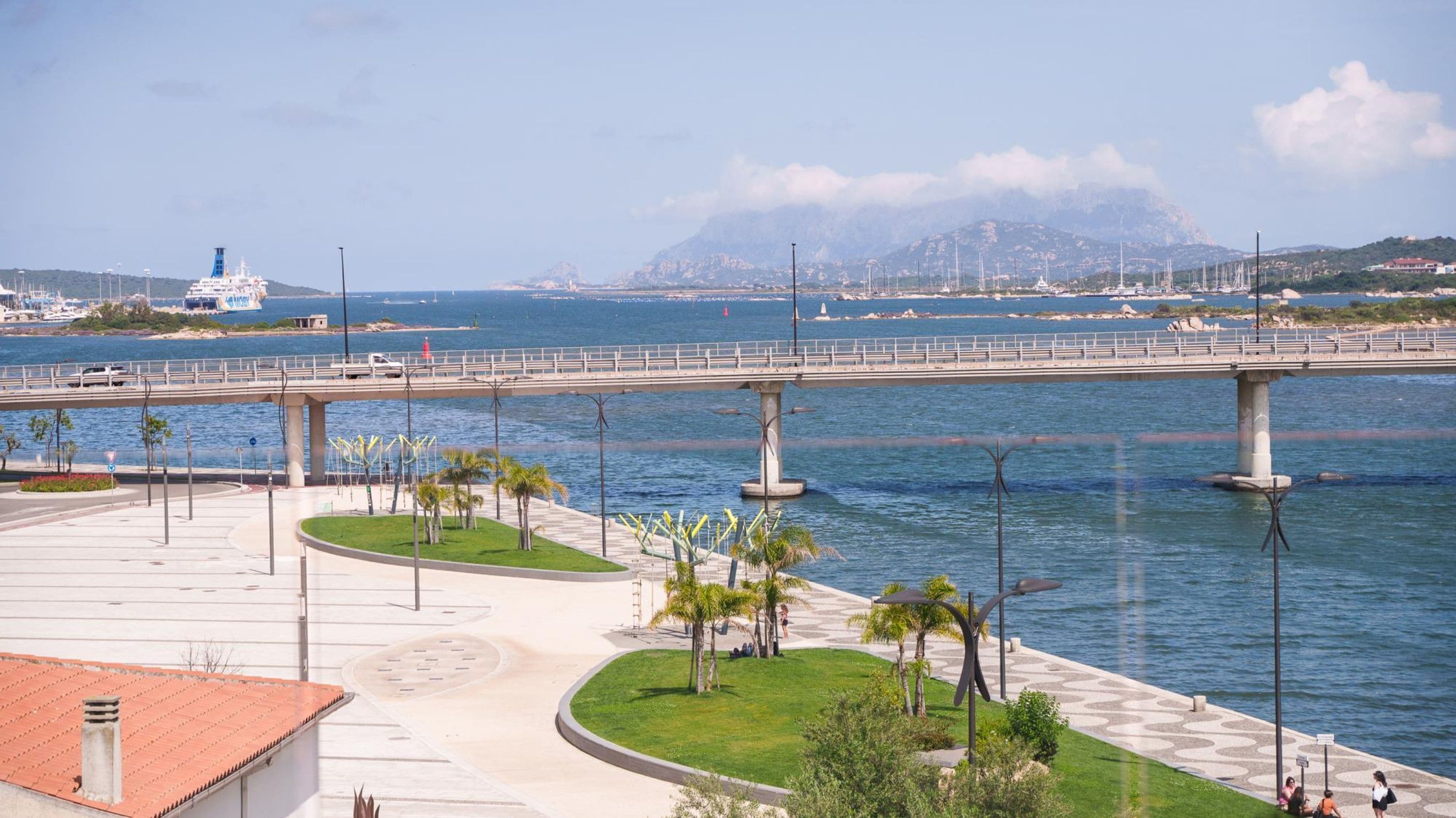 Um calçadão à beira-mar com uma ponte, palmeiras e vista para o mar e montanhas ao longe.