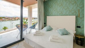 Quarto de hotel com uma cama grande, duas almofadas, vista para a água e varanda com cadeiras.