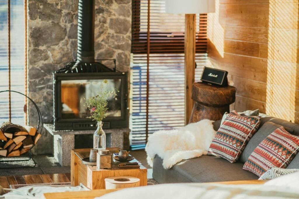 Uma sala de estar acolhedora com parede de pedra, lareira, sofá com almofadas estampadas e uma pequena mesa de madeira com um vaso.