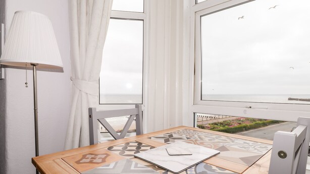 Quarto com uma mesa de madeira, uma luminária e vista para o mar através de amplas janelas.