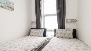 Quarto com duas camas de solteiro, cada uma com cabeceira e travesseiro estampado com motivos florais. Há uma janela com cortinas, um luminária fixada na parede e um quadro na parede.