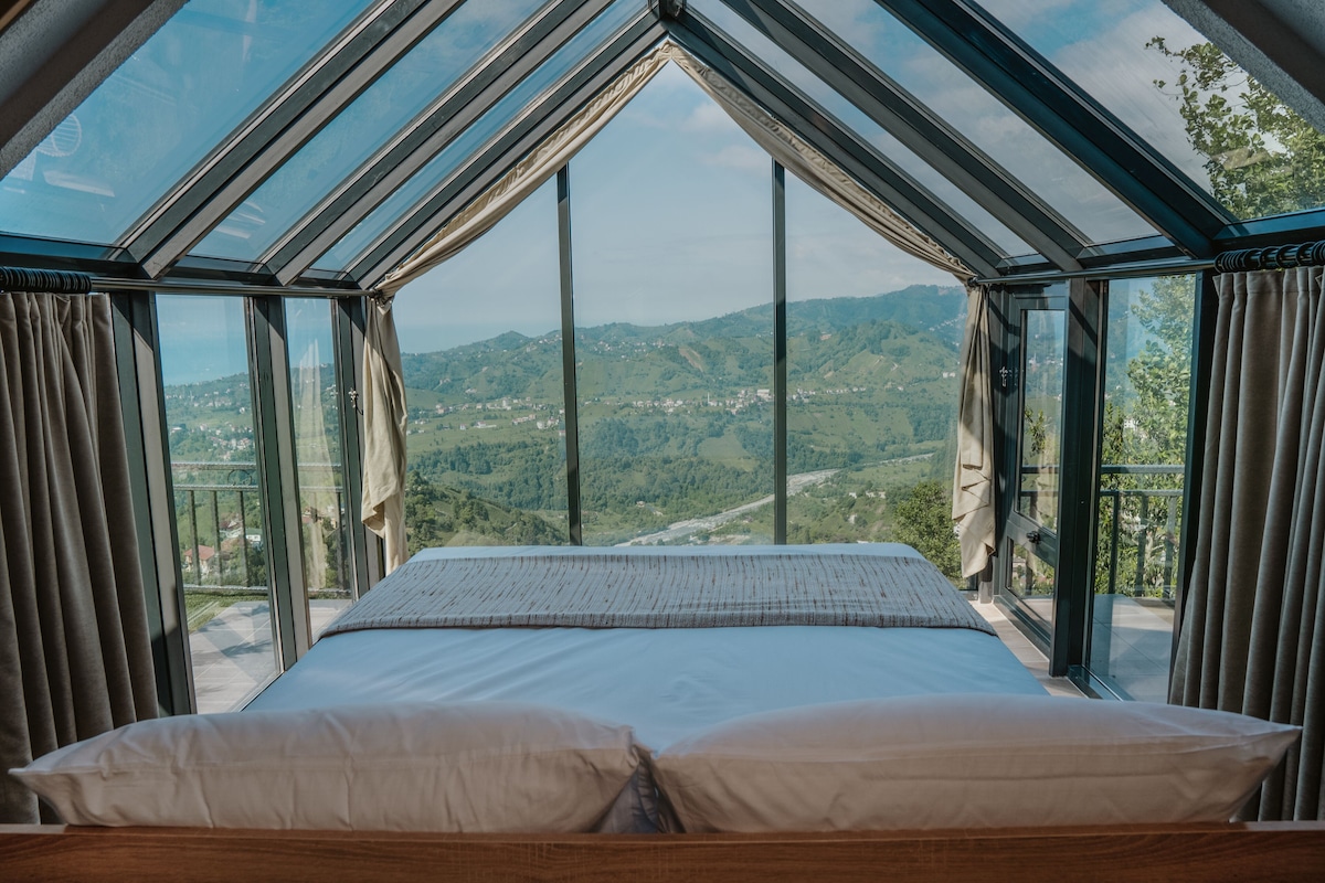 Uma cama com vista para uma paisagem montanhosa através de grandes janelas de vidro.