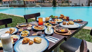 Uma mesa de jantar com diversos itens de café da manhã e um copo de suco de laranja, com vista para a piscina.