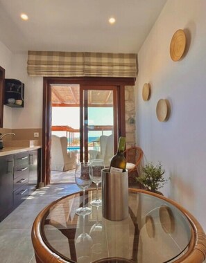 Cozinha moderna com mesa de vidro, copos de vinho e uma garrafa de vinho. Há vista do exterior por uma porta de vidro.
