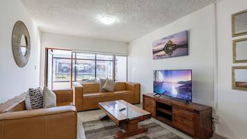 Uma sala de estar com um sofá, uma mesa de centro, uma TV sobre um suporte de madeira e uma janela grande.