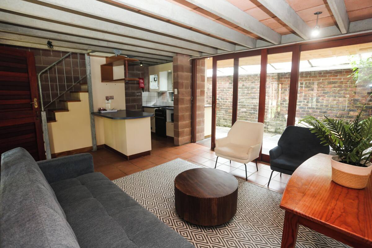 Uma sala de estar com sofá, uma mesa de centro redonda, uma mesa de jantar com cadeiras, uma escada e uma área de cozinha.
