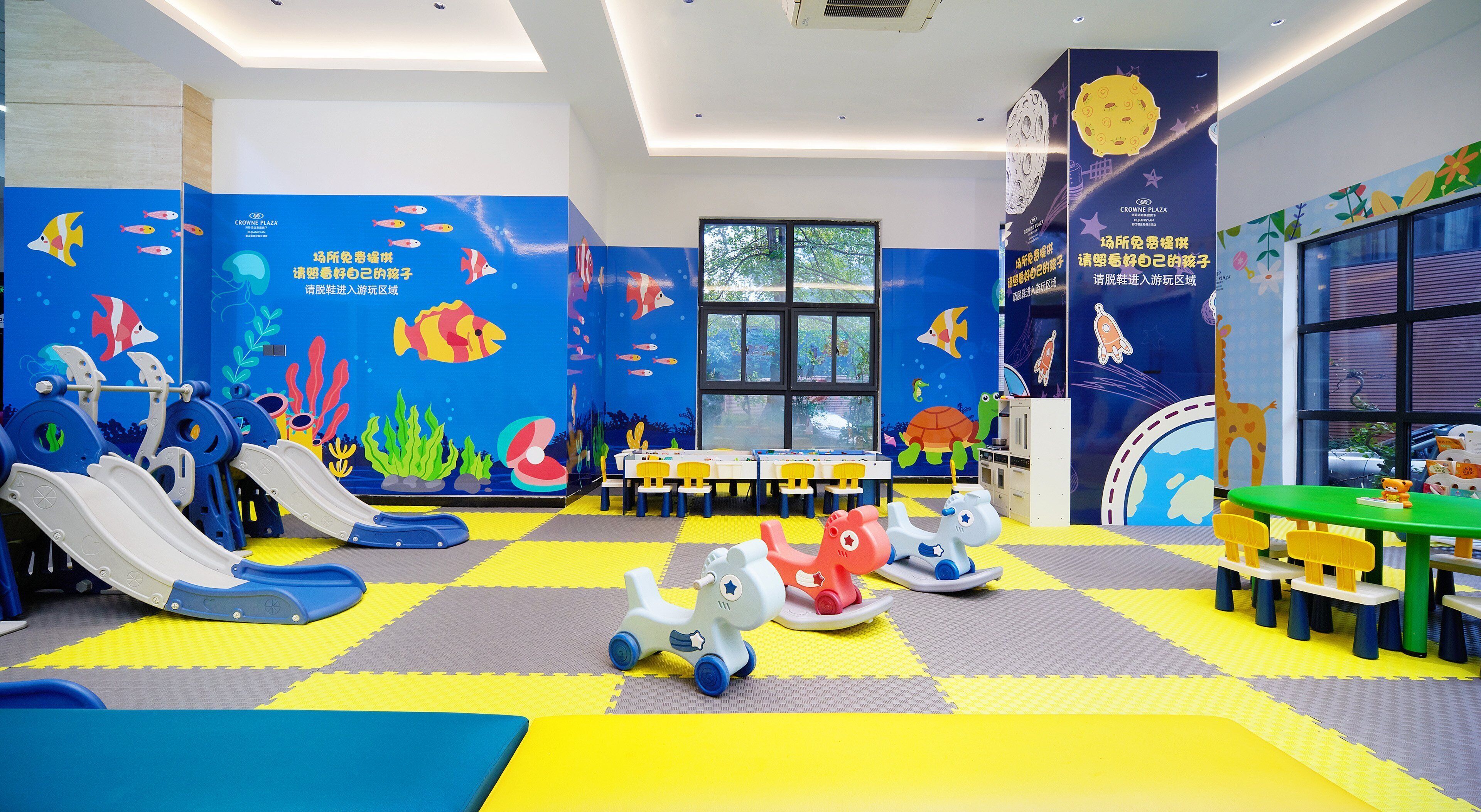 Espaço infantil com escorregadores, mesas e painéis coloridos nas paredes.