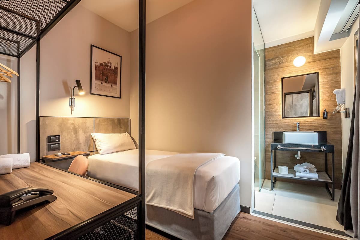 Quarto de hotel com duas camas, uma mesa com telefone, um quadro na parede e um banheiro com pia e espelho.