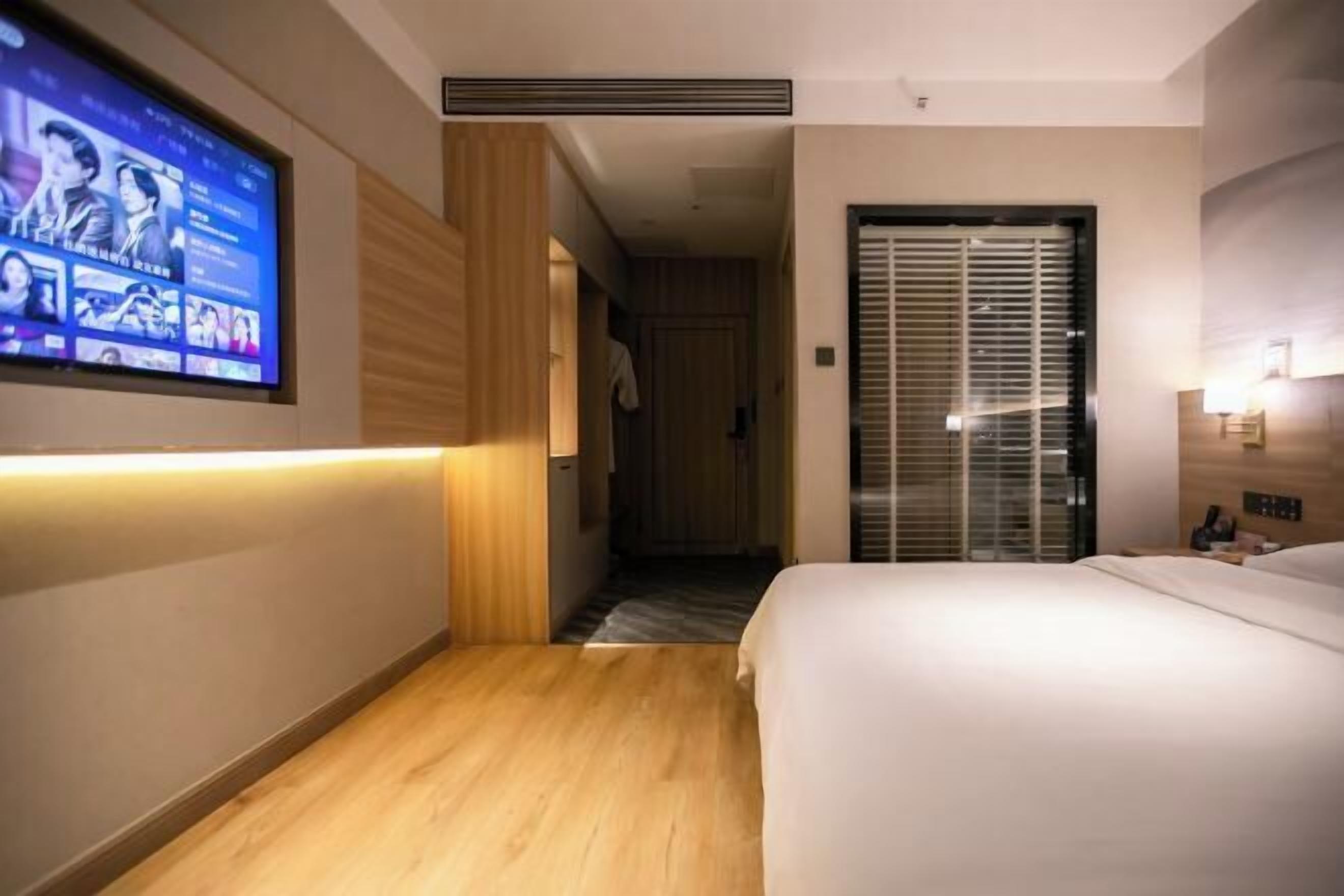 Quarto de hotel com cama, televisão fixada na parede, porta de madeira e janela com persianas.
