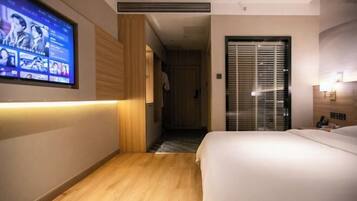 Quarto de hotel com cama, televisão fixada na parede, porta de madeira e janela com persianas.