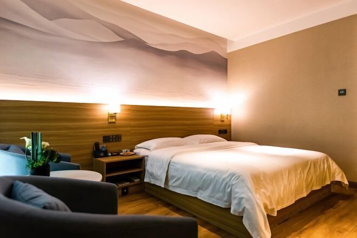 Quarto com cabeceira de madeira, cama com roupa de cama branca, mesa de cabeceira com abajur, uma planta pequena e uma cadeira.