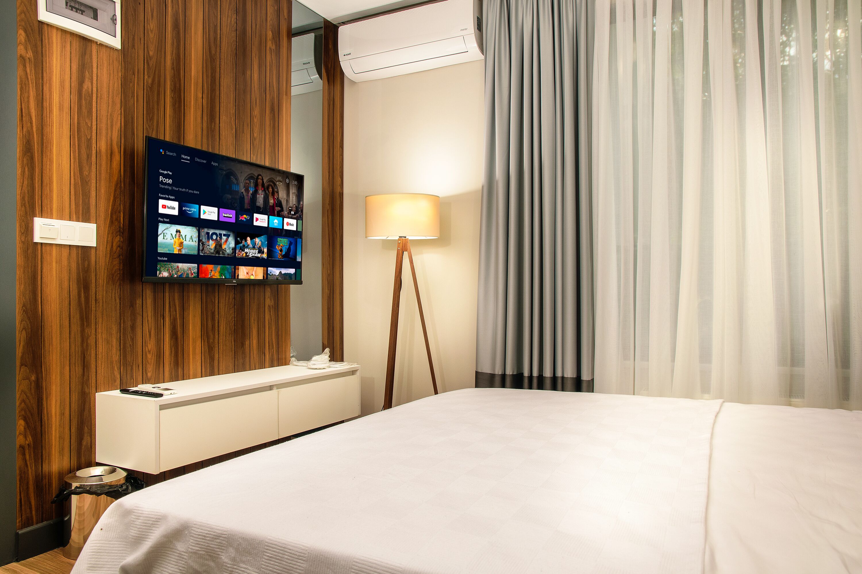 Quarto de hotel com uma cama grande, uma televisão fixada numa parede de madeira, uma mesa de cabeceira com abajur e uma janela com cortinas leves.