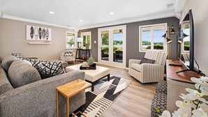 Uma sala de estar moderna com um sofá cinzento, uma poltrona riscada e uma mesa de centro de madeira. Há grandes janelas com vista para o verde lá fora.