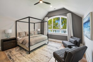 Um quarto com cama de dossel, mesa de cabeceira, cadeira e uma janela grande com cortinas.