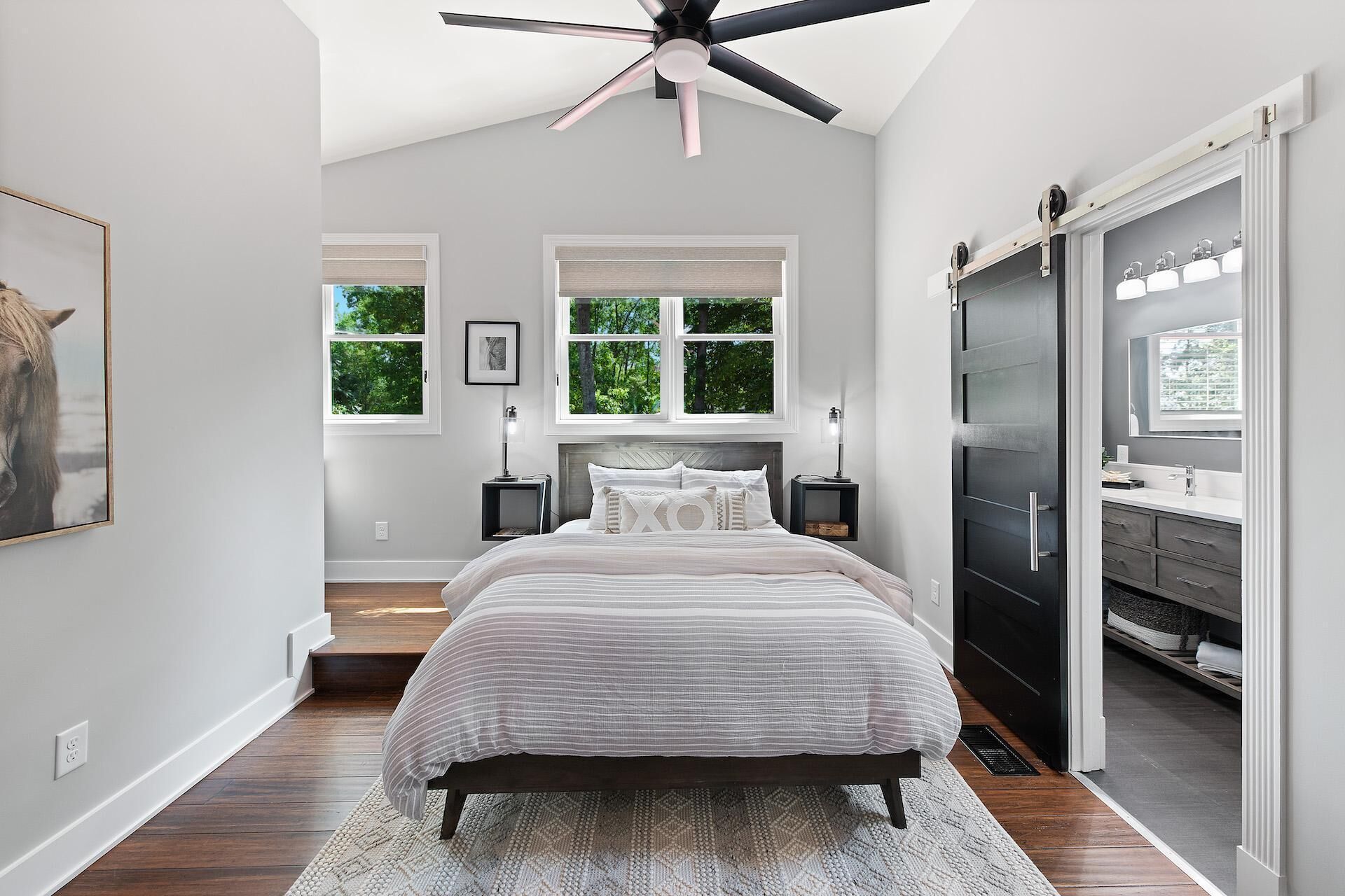 Quarto com cama, mesas de cabeceira, ventoinha no teto e vista para o exterior pelas janelas.