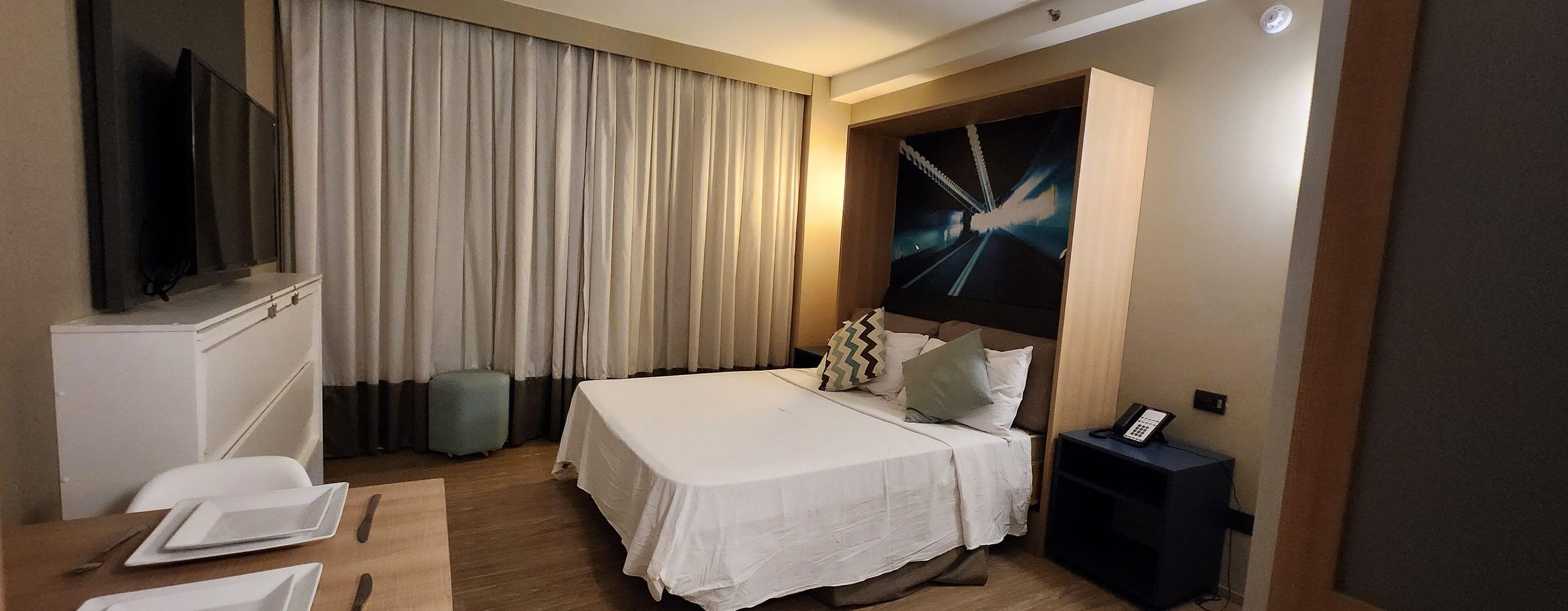 Quarto de hotel com cama, escrivaninha com cadeira, telefone, televisão e uma janela grande com cortinas.
