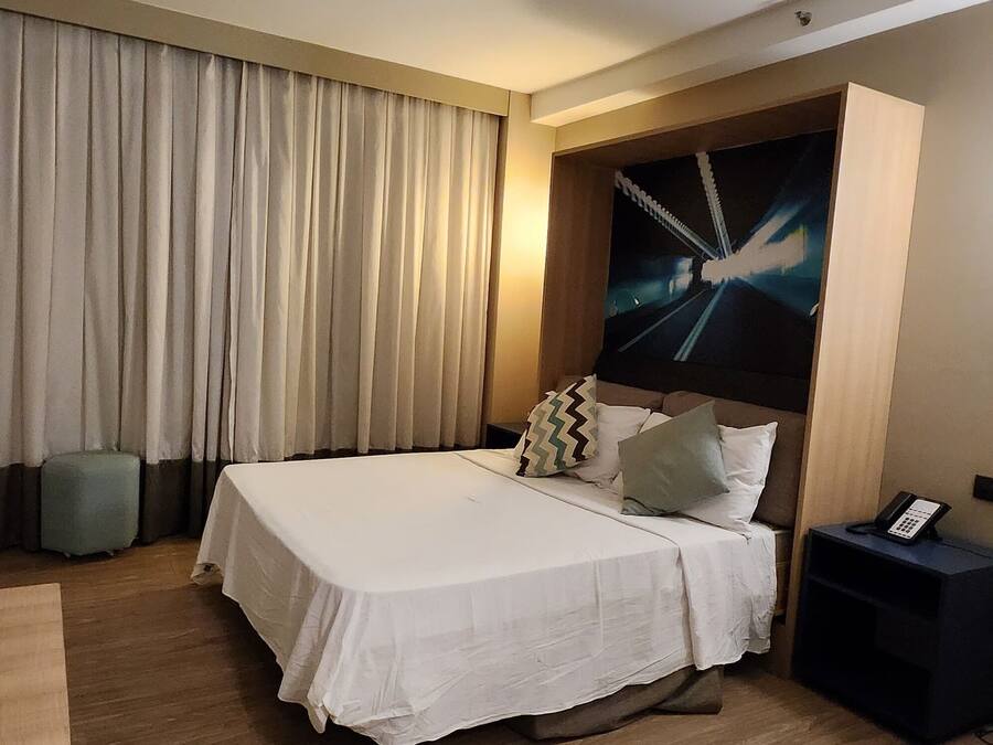 Quarto de hotel com cama, escrivaninha com cadeira, telefone, televisão e uma janela grande com cortinas.