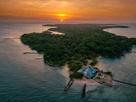 Vista aérea de uma ilha tropical com vegetação densa, um pequeno cais e um edifício com telhado azul.