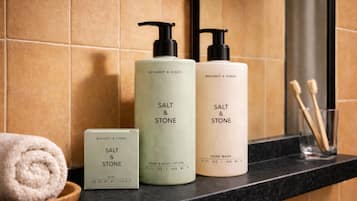 Uma prateleira no banheiro com três frascos de sabonete Bergamot & Hinoki Salt & Stone, uma toalha enrolada e um copo com escovas de dentes.