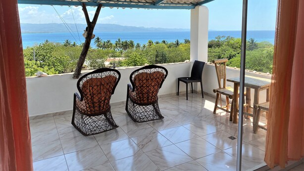 Uma varanda com duas cadeiras de vime, uma mesa pequena e vista para o oceano e vegetação.
