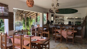Uma zona de refeições rústica com mesas e cadeiras de madeira, um candeeiro pendente e um espaço bar com prateleiras.