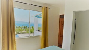 Quarto com uma cama, janela com cortinas e vista para o mar e vegetação.