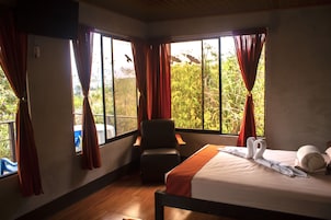 Quarto de hotel com cama, uma cadeira e amplas janelas com cortinas.