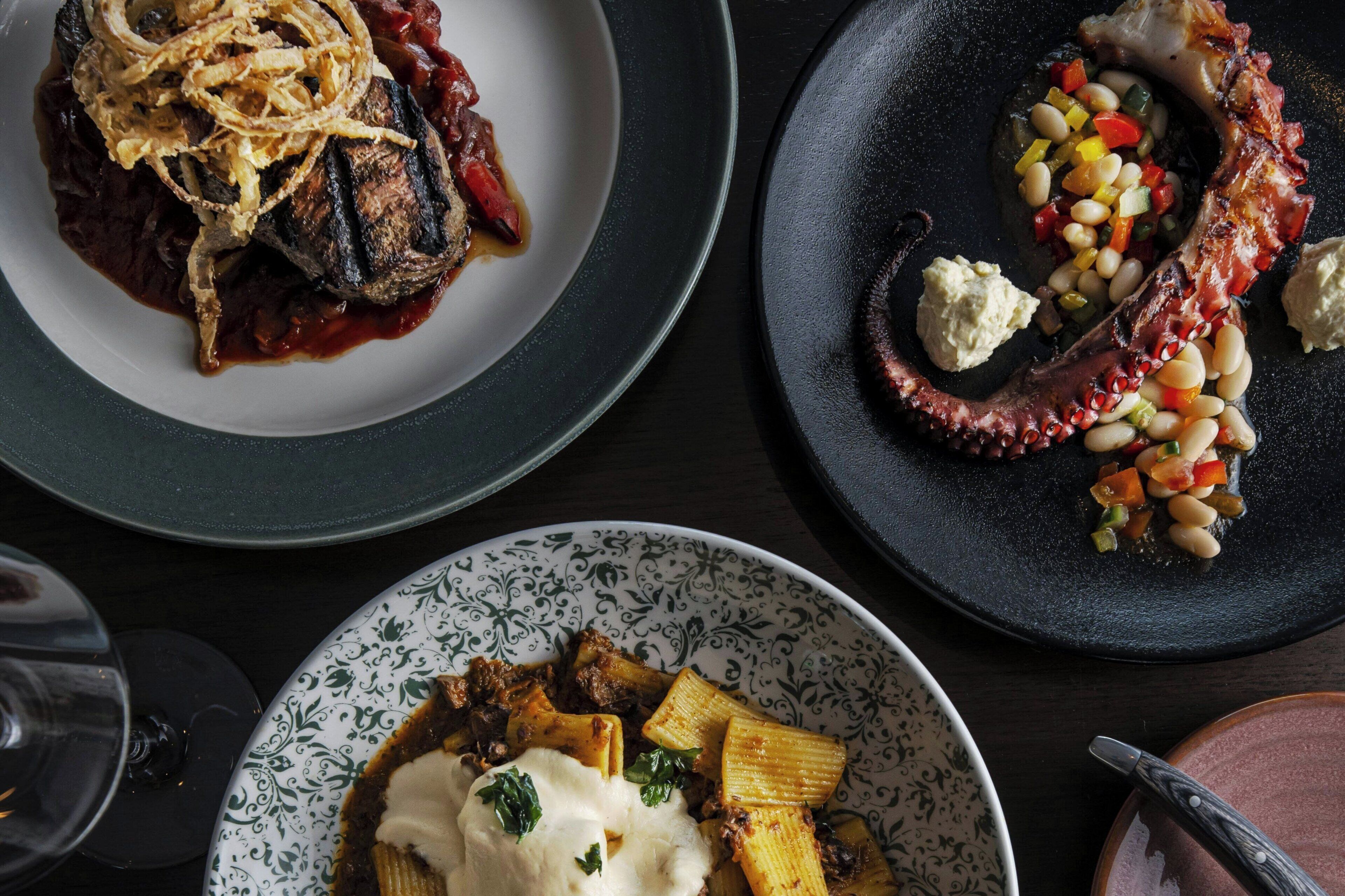 Uma mesa com três pratos de comida, incluindo bife com cebolas, polvo com legumes e massa com molho cremoso.
