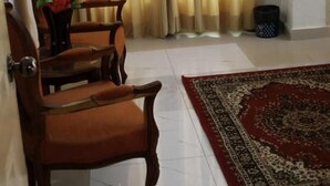 Uma sala de estar com um tapete estampado vermelho, cadeiras de madeira, uma mesa com um vaso de flores, cortinas bege e um ar-condicionado fixado na parede.