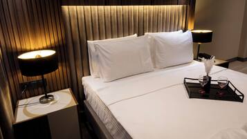 Quarto de hotel com uma cama, duas luminárias de cabeceira, um suporte com copos e um vaso, e um painel de cabeceira de madeira.
