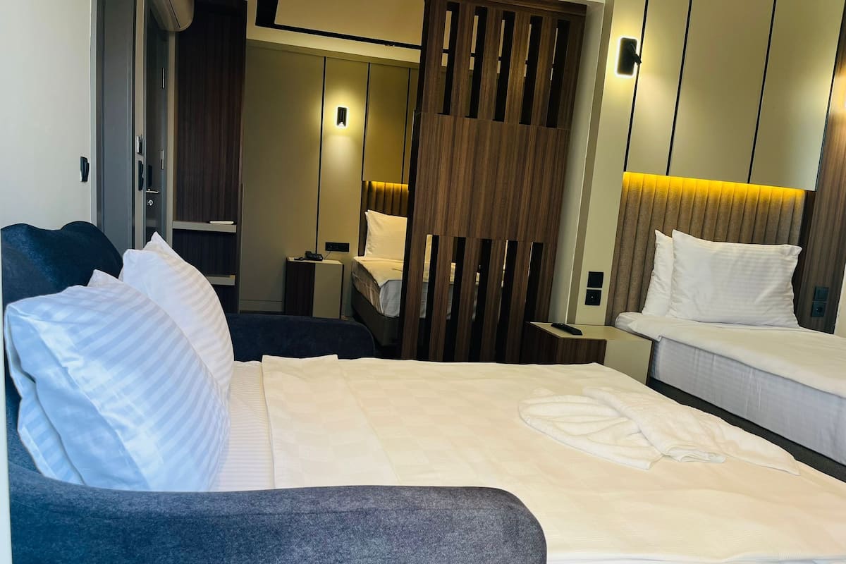 Quarto de hotel moderno com uma cama grande, um sofá e uma escrivaninha com uma cadeira. Há uma divisória de madeira e uma pequena mesa com um abajur.