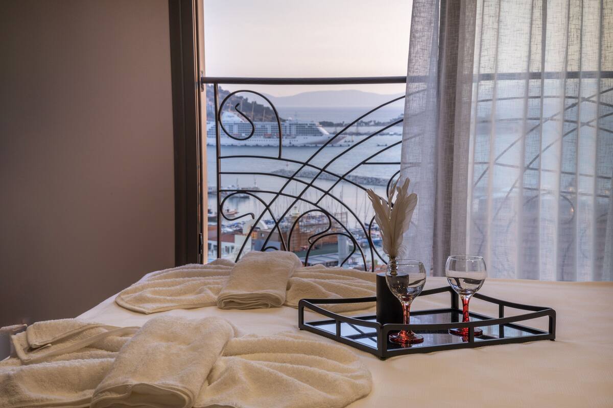 Uma varanda com guarda-corpo de metal, uma mesa com toalhas e um copo de vinho, e vista para a cidade e montanhas.