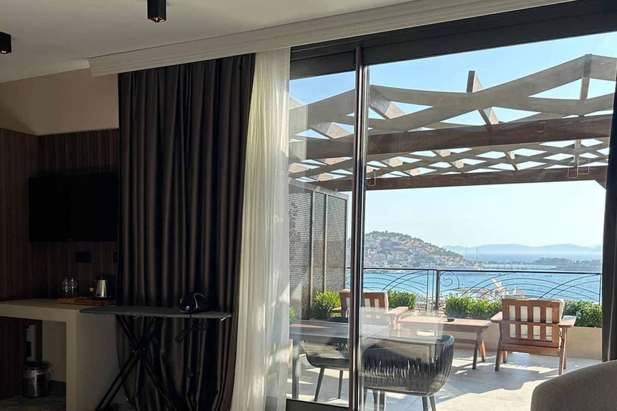 Quarto de hotel com uma cama, uma mesa, uma cadeira e uma varanda com vista para o mar e montanhas.
